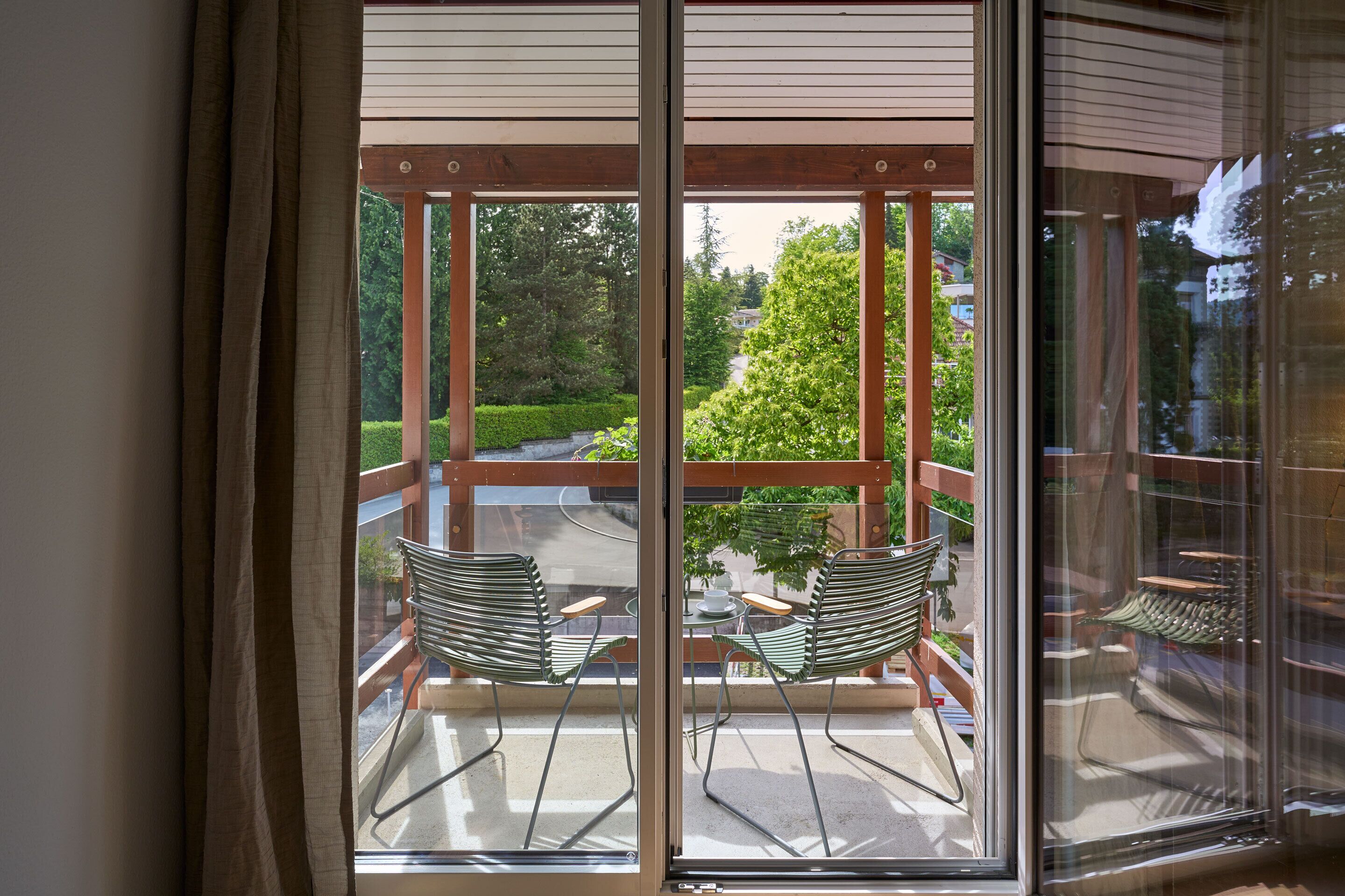 Uma varanda com duas cadeiras e uma mesa, com vista para um jardim e uma rua.