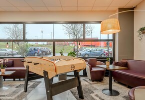 Quarto com mesa de pebolim, sofá, mesa com abajur e vista para carros estacionados lá fora.