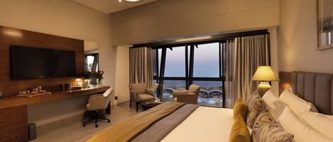 Quarto de hotel com uma cama grande, uma escrivaninha com televisão de tela plana, uma cadeira, um abajur e vista para a cidade.