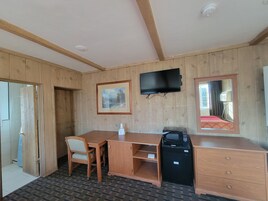 Quarto com escrivaninha de madeira, cadeira e micro-ondas preto. Há uma televisão montada na parede e uma janela com cortinas.