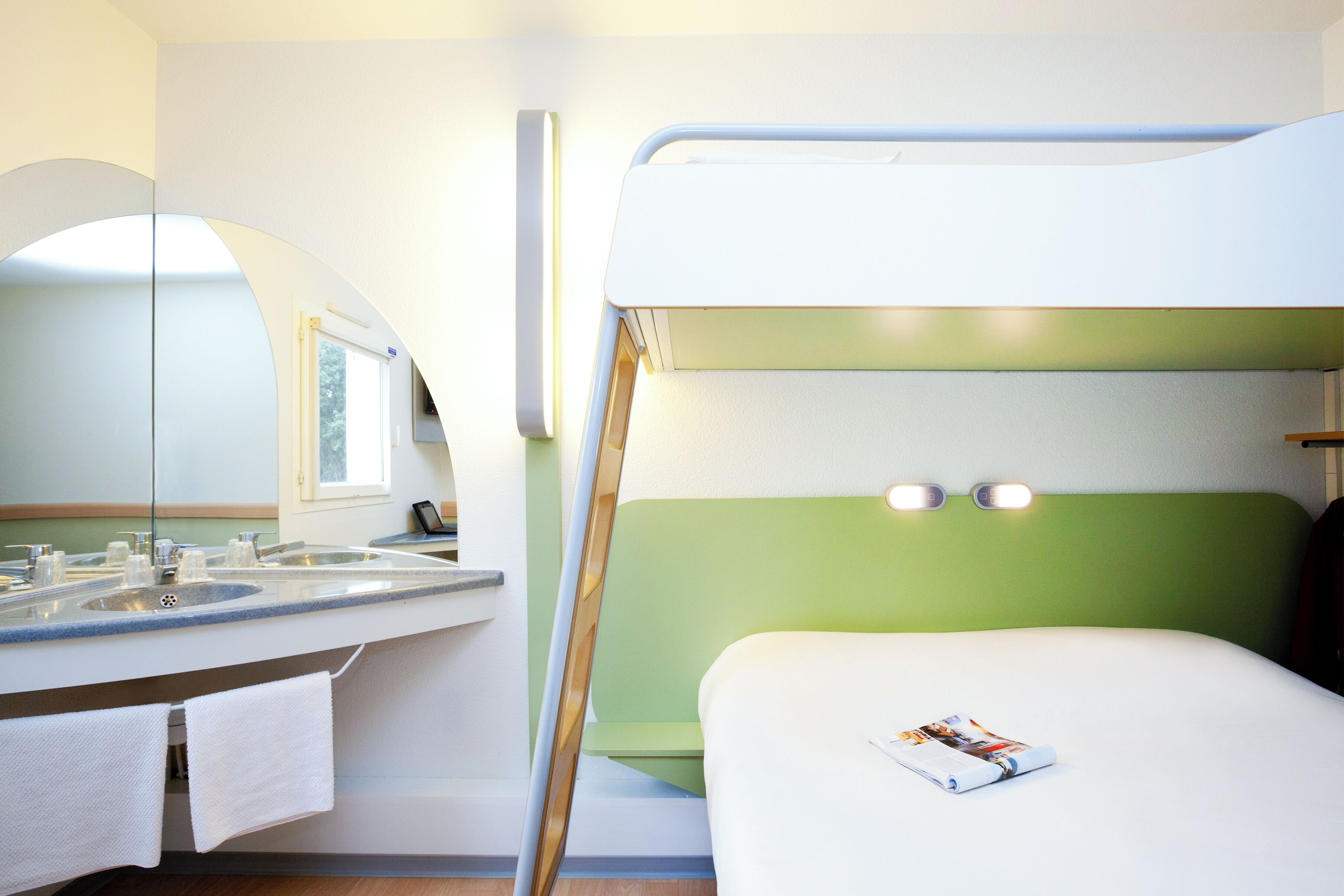 Quarto de hotel compacto com uma cama beliche, uma pia com espelho e uma pequena mesa de cabeceira.