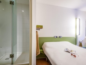 Quarto de hotel com box de vidro, cama com cabeceira verde, mesa de cabeceira com um livro e óculos de sol, e luminária fixada na parede.