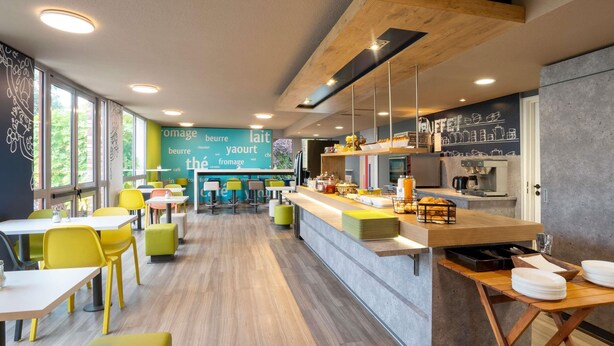 Um café moderno com piso de madeira, mesas amarelas e brancas e um balcão central com alimentos.