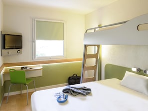 Quarto de hotel com beliche, escrivaninha com cadeira, televisão e janela.