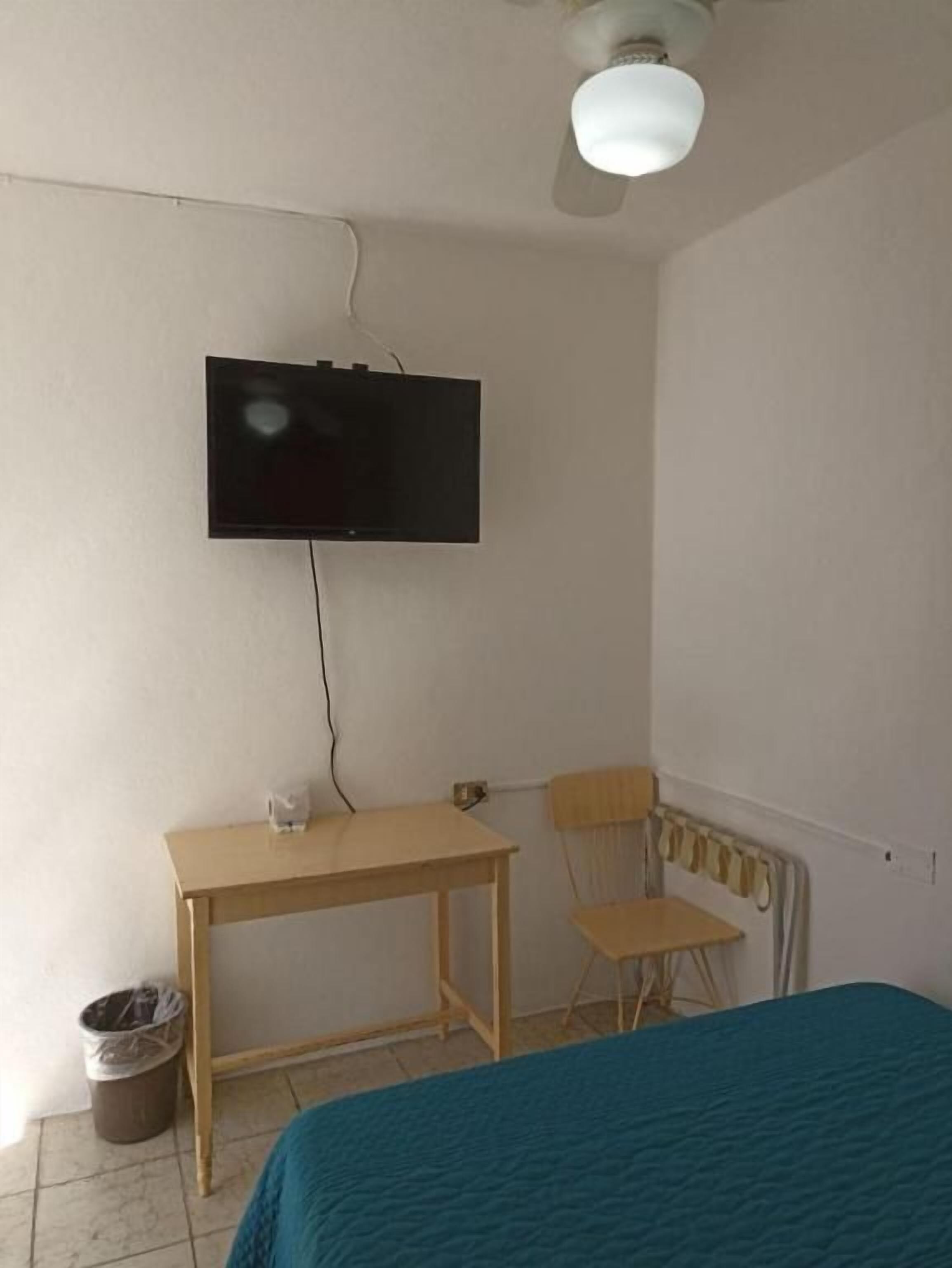 Quarto com cama, escrivaninha, cadeira, televisão fixada na parede e ventoinha no teto.