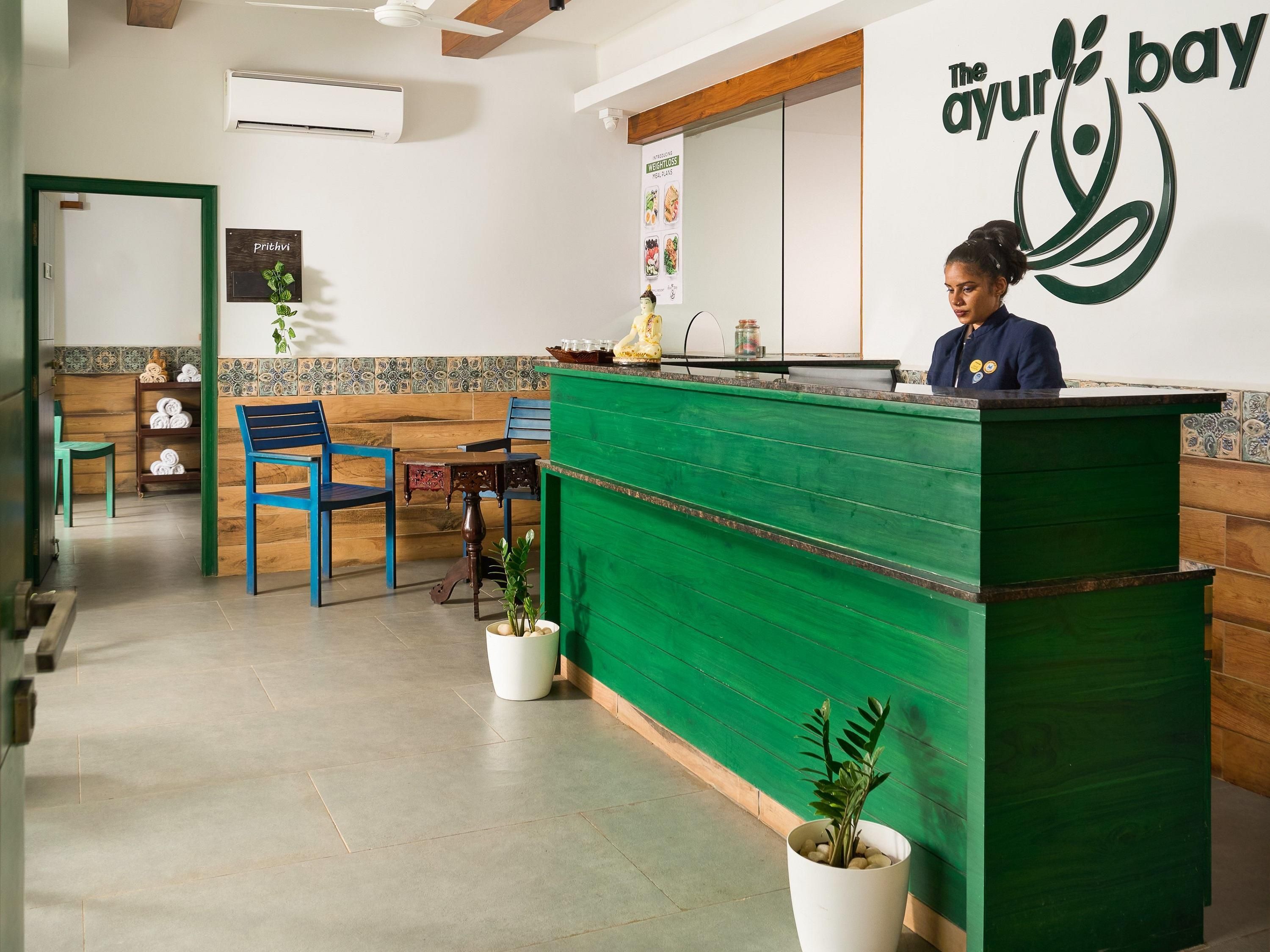 Uma área de recepção com balcão verde, um banco de madeira e parede com o texto 'The ayur bay'.