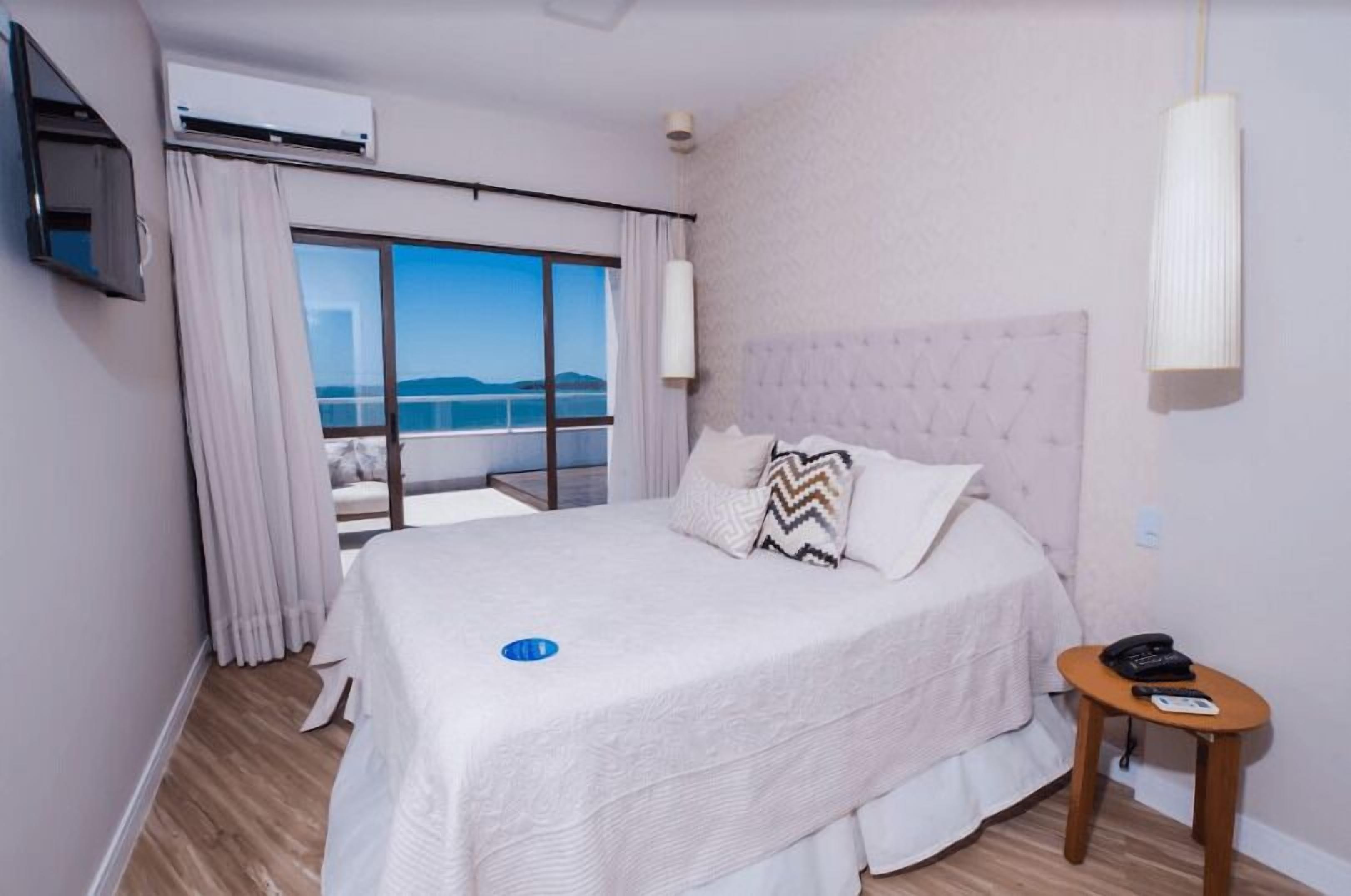 Quarto de hotel com uma cama grande, uma mesa de cabeceira com telefone e vista para o mar através de uma janela ampla.