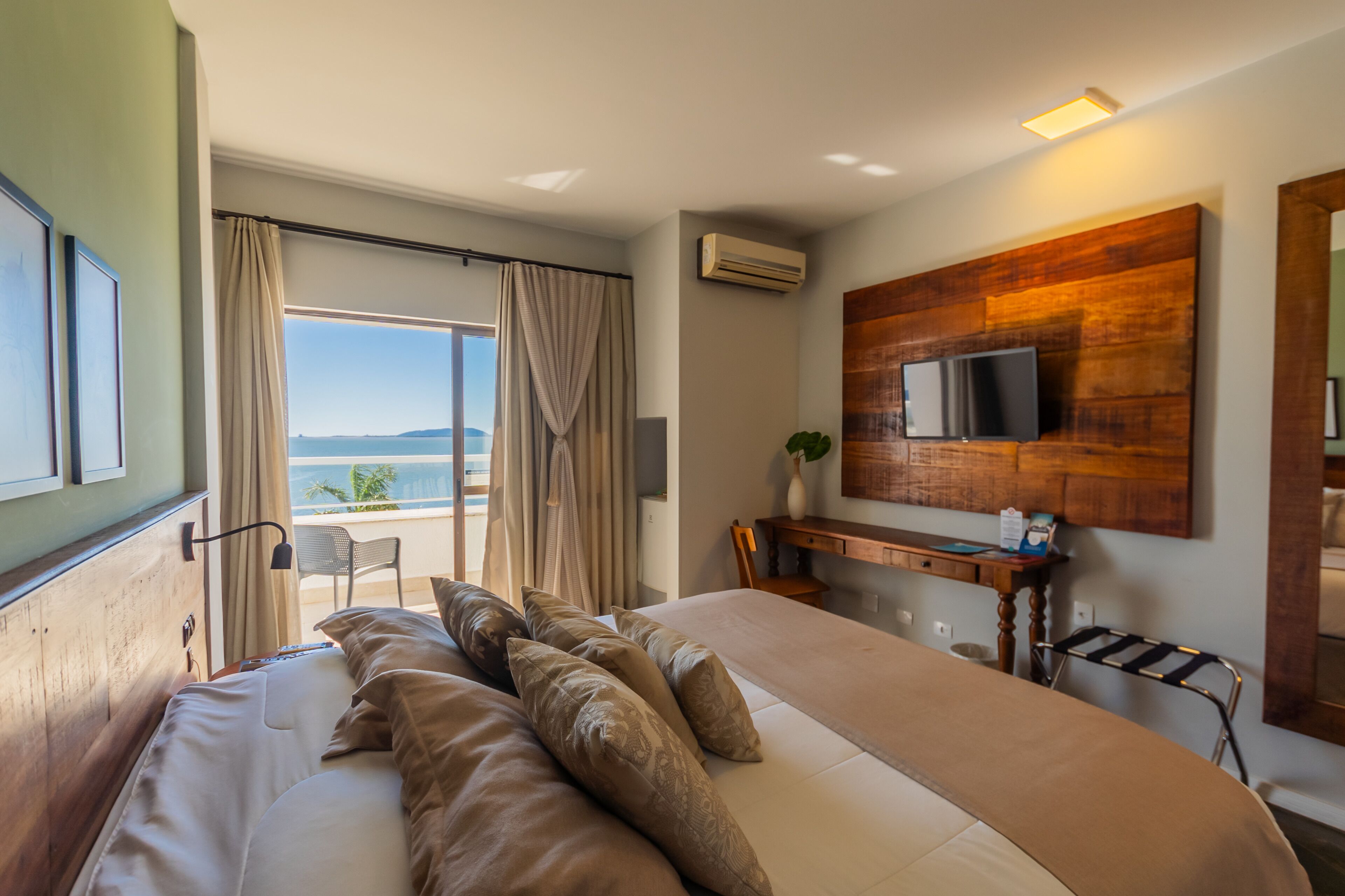 Quarto de hotel com uma cama grande, uma escrivaninha, uma TV fixada num painel de madeira e vista para o mar.