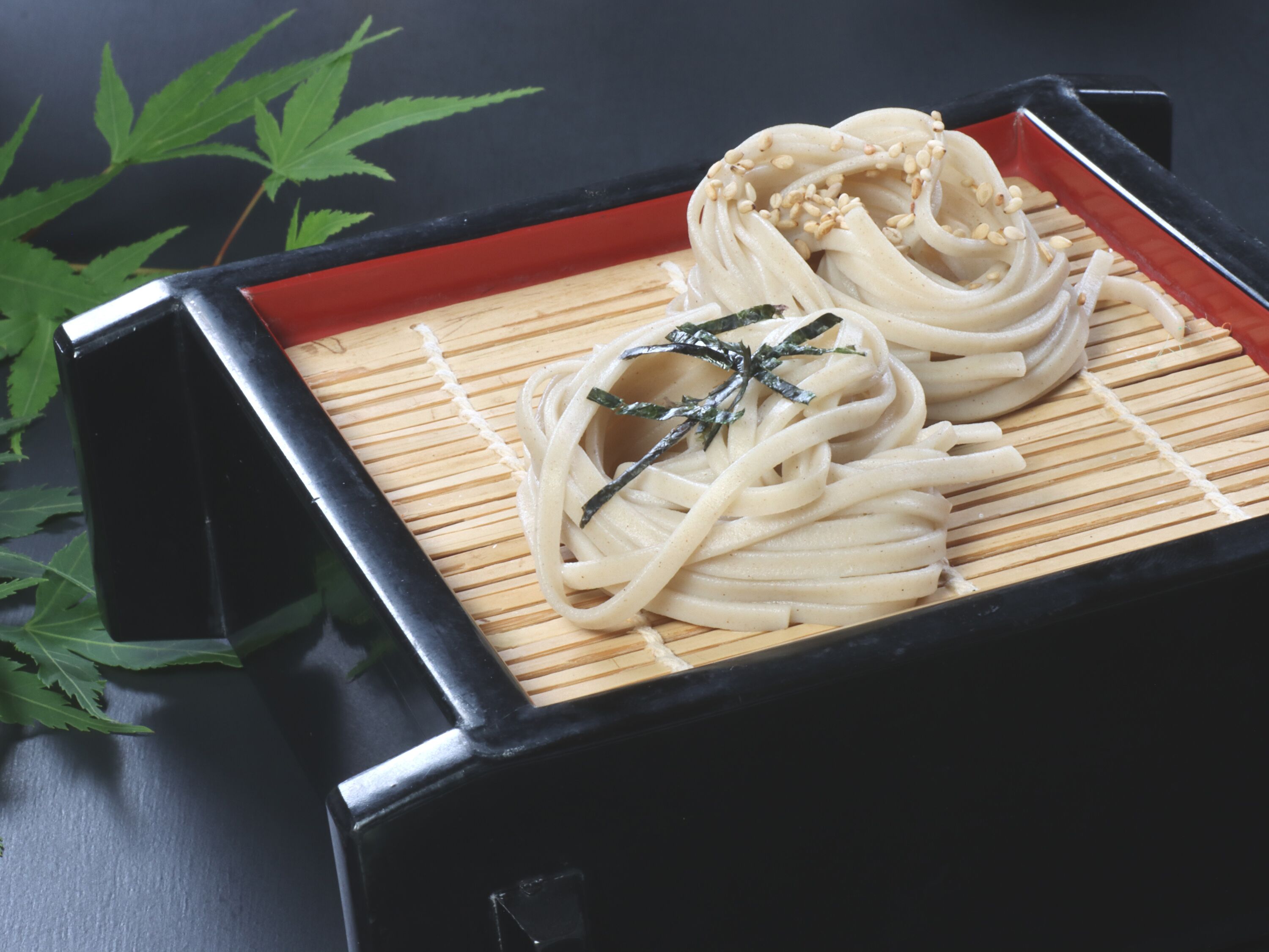 Uma travessa com macarrão branco, sementes de gergelim e folhas verdes sobre um tapete de bambu.