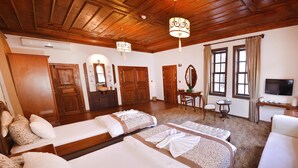Quarto de hotel com tetos de madeira, duas camas com roupa de cama branca, uma mesa pequena, uma cadeira, uma televisão e uma janela com cortinas.