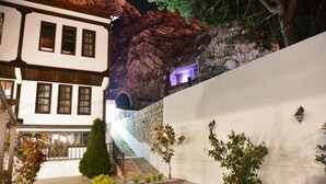 Um edifício com escadaria de pedra que leva a uma entrada iluminada, rodeado por vegetação e uma parede branca.