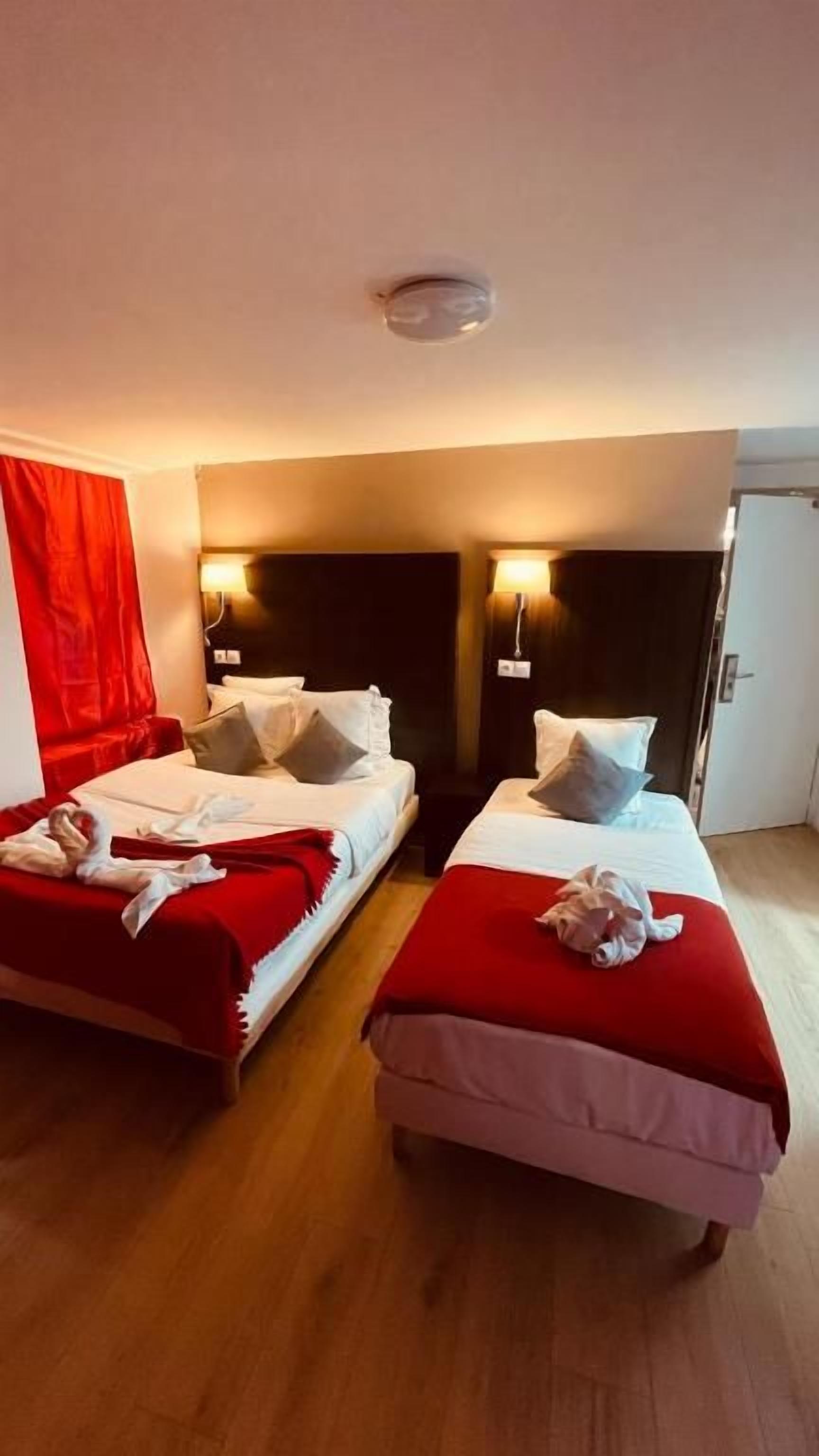 Quarto de hotel com duas camas, cada uma com colcha e almofadas vermelhas, e uma cabeceira escura.
