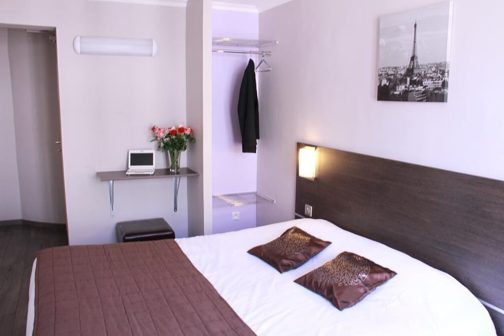 Quarto de hotel com cama, mesa de cabeceira, abajur, quadro da Torre Eiffel e um cabideiro.