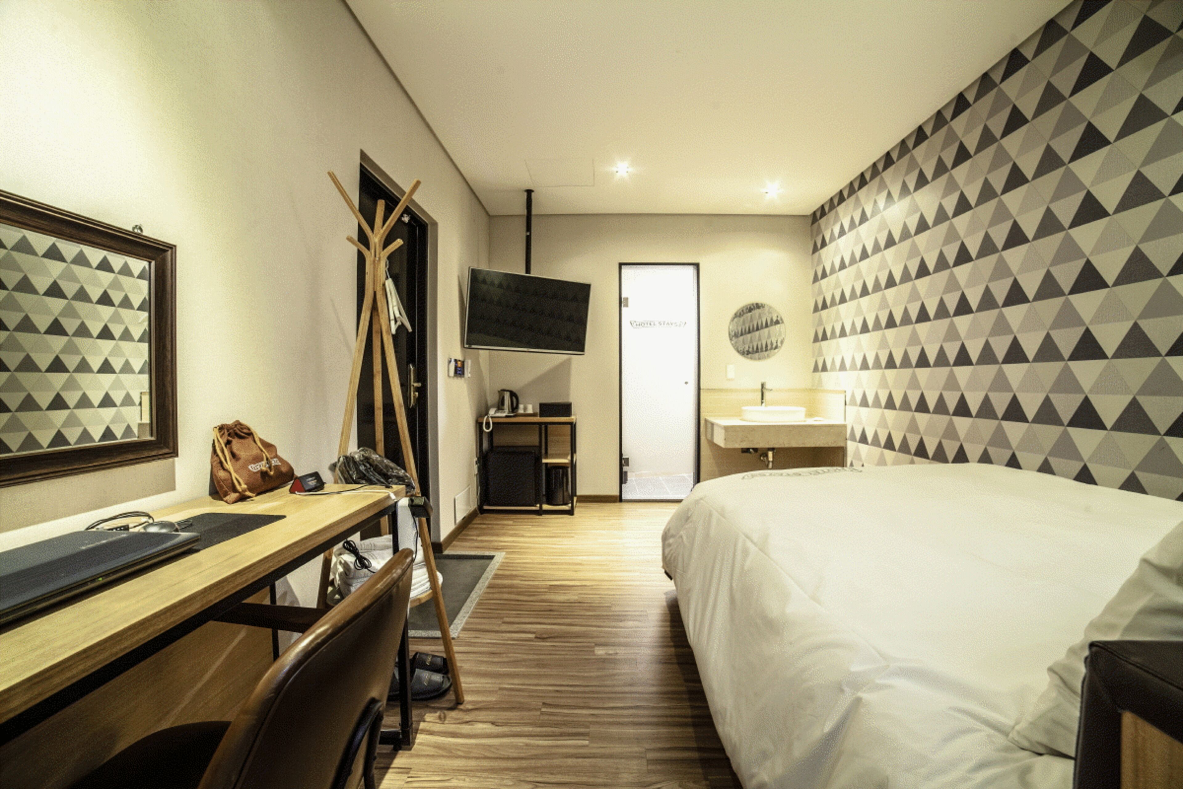Quarto de hotel com cama, escrivaninha, cadeira, televisão e parede estampada.