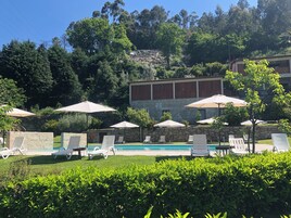 Uma área de piscina com espreguiçadeiras e guarda-sóis, cercada por vegetação e um edifício de pedra.
