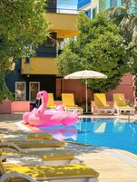 Uma área de piscina com boia de flamingo rosa, espreguiçadeiras amarelas e um guarda-sol branco.