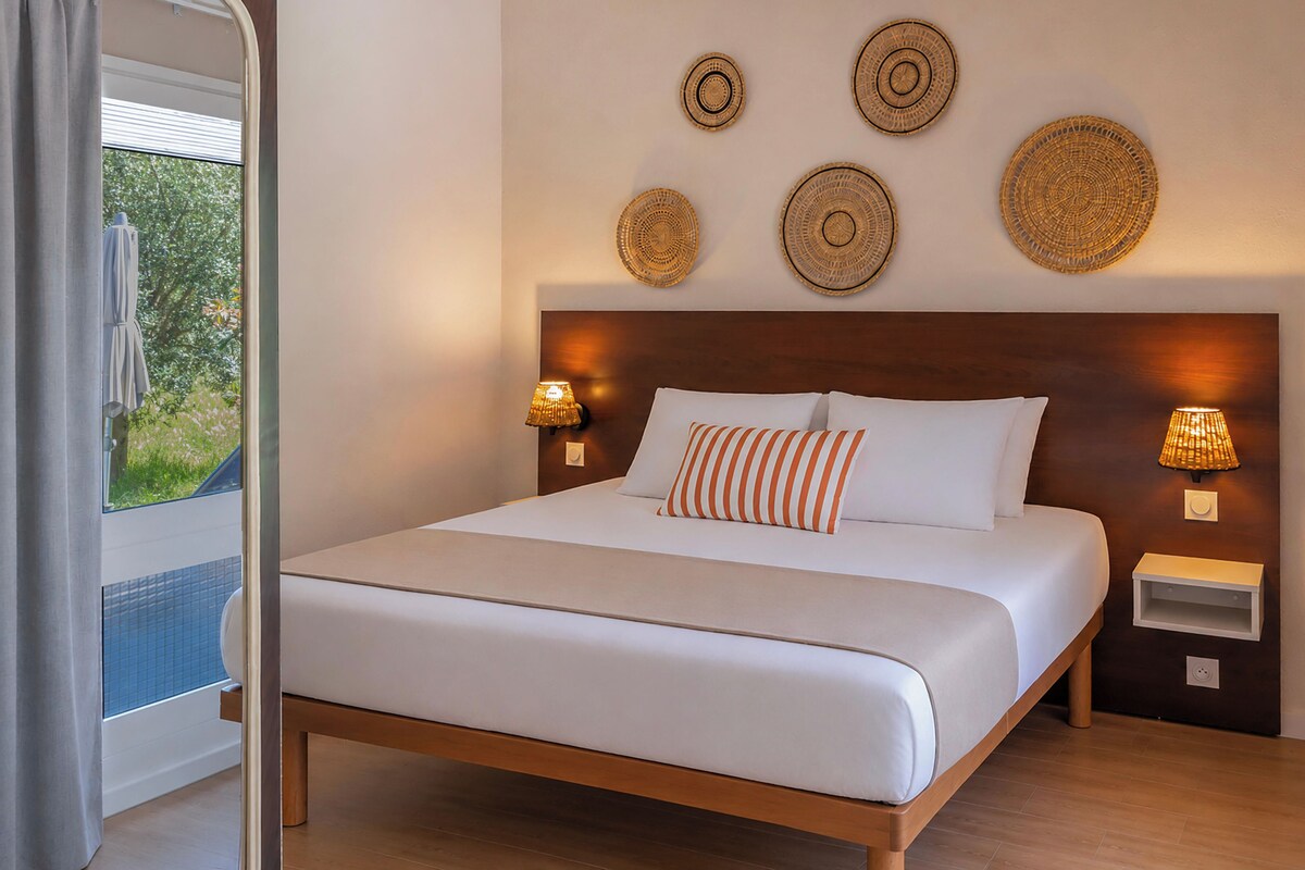 Quarto de hotel com cama, abajures, luminária na parede e vista para o verde lá fora.