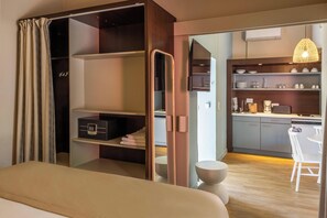 Quarto de hotel com cama, guarda-roupa, espelho, mesinha, cadeira e cozinha equipada com pia e mesa de jantar.