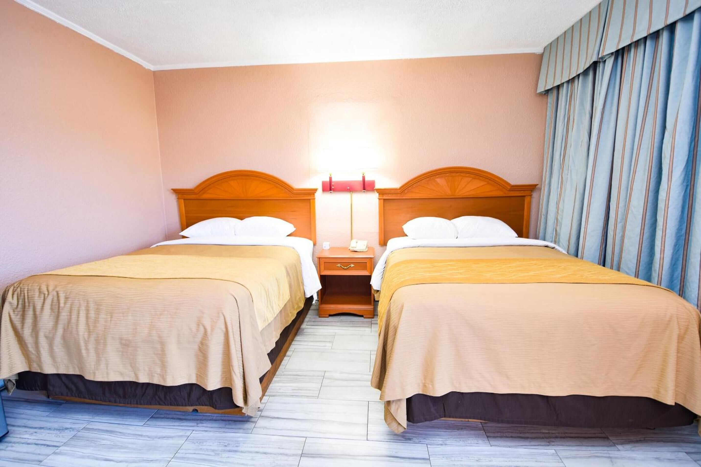 Quarto de hotel com duas camas de solteiro, uma mesa de cabeceira com luminária e uma janela com cortinas.