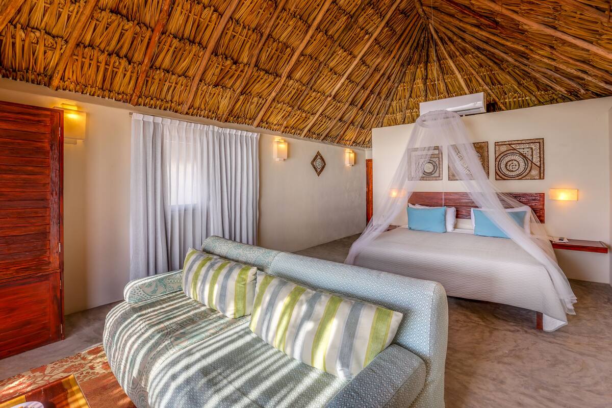 Quarto com cama, sofá, telhado de palha e luminárias fixadas na parede.