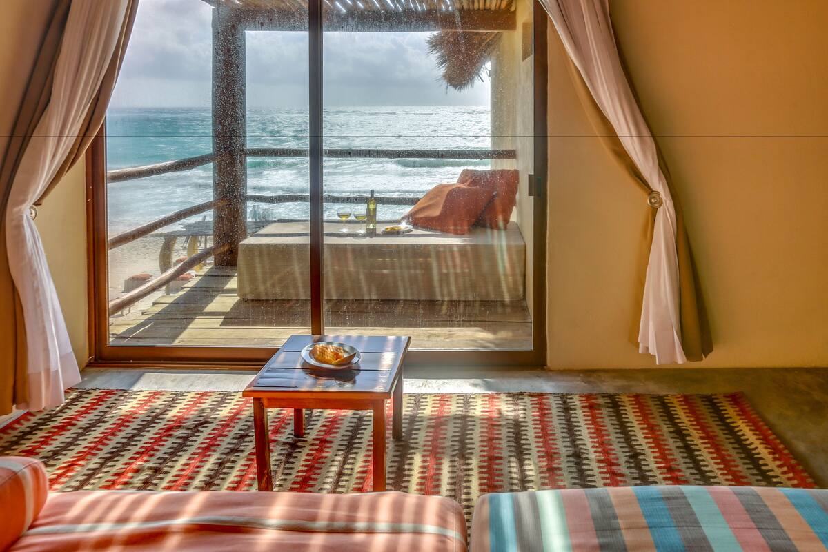 Quarto com uma grande porta de vidro deslizante que dá para uma varanda com vista para o oceano. O quarto possui um tapete estampado, uma pequena mesa com uma tigela e uma garrafa, e uma área de estar acolchoada.