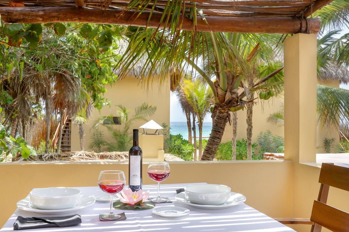 Mesa posta para dois, com uma garrafa de vinho e copos, com vista para uma praia com palmeiras e o oceano.