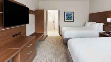 Quarto de hotel com televisão de tela plana fixada em um armário de madeira, cama com roupa de cama branca, pequena mesa de cabeceira com abajur e uma obra de arte abstrata emoldurada na parede.