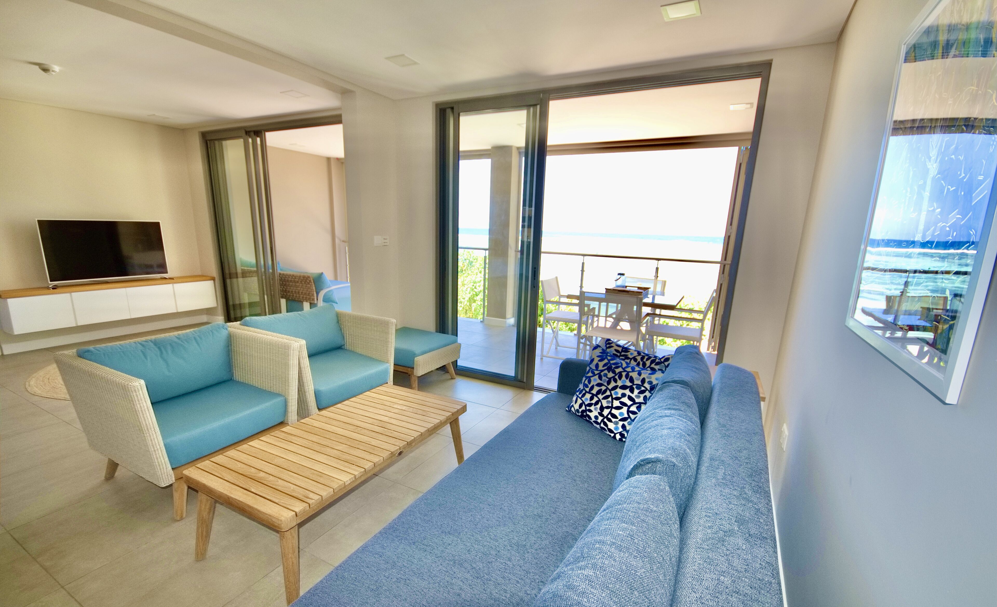 Uma sala de estar moderna com um sofá, duas poltronas, uma mesa de centro e uma televisão de tela plana em um armário branco. Grandes portas de vidro dão acesso a uma varanda com vista para o mar.