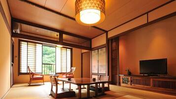 Quarto tradicional japonês com tatames, uma mesa baixa e cadeiras. Há uma televisão sobre um suporte de madeira, e uma grande janela com vista para o verde.