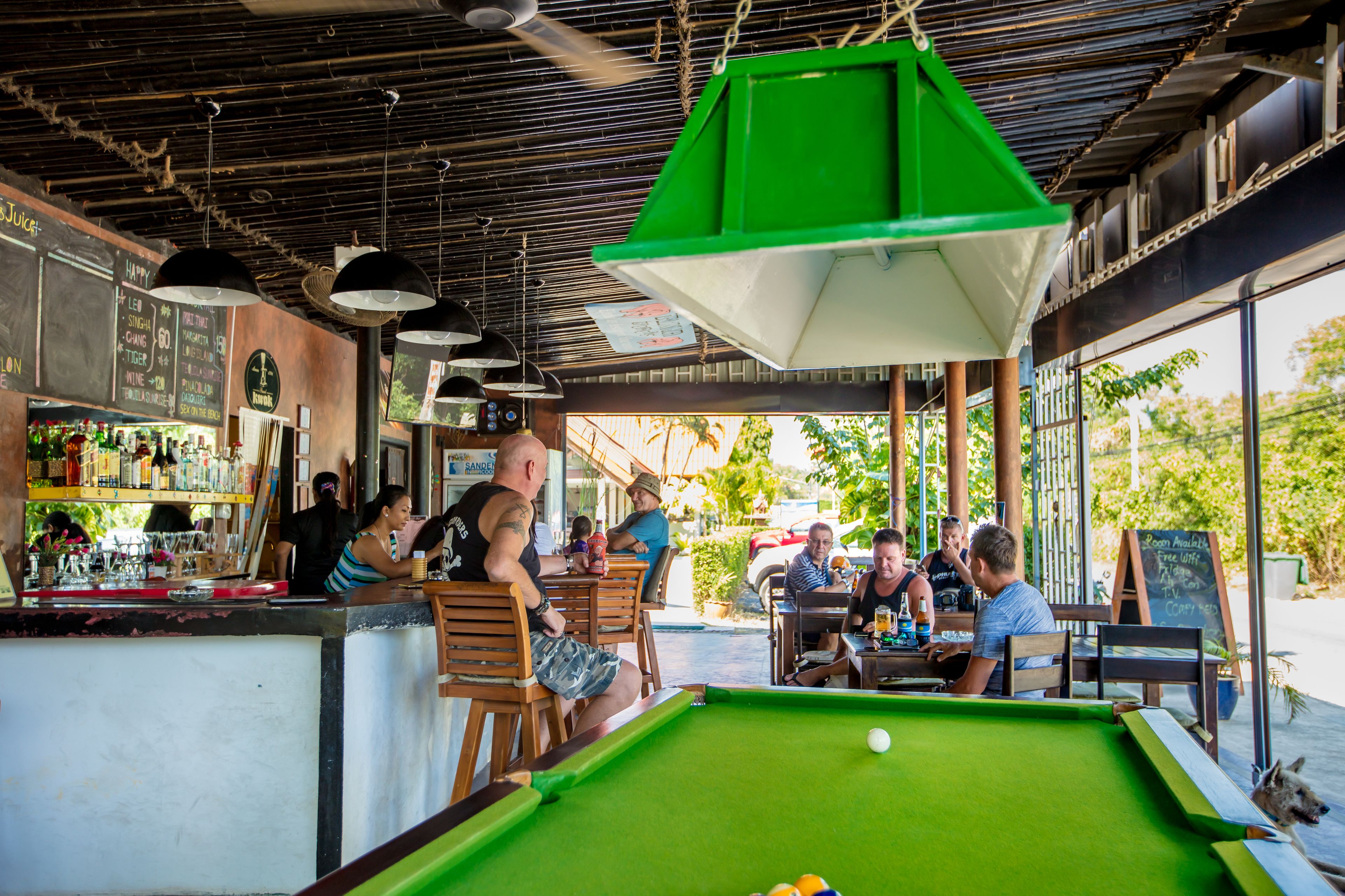 Uma mesa de bilhar em uma área de bar, com pessoas ao redor.