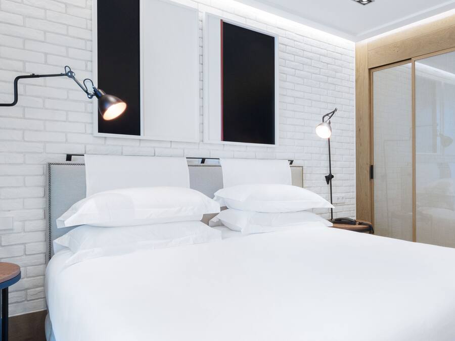 Quarto de hotel moderno com parede de tijolos brancos, uma cama grande com roupa de cama branca, duas luminárias pretas fixadas na parede e um guarda-roupa de madeira.