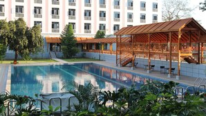 Piscina com deck de madeira e espreguiçadeiras, cercada por vegetação e um edifício alto ao fundo.