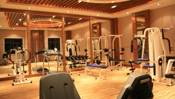 Academia com diversos equipamentos de exercício, piso de madeira e um espelho grande na parede.