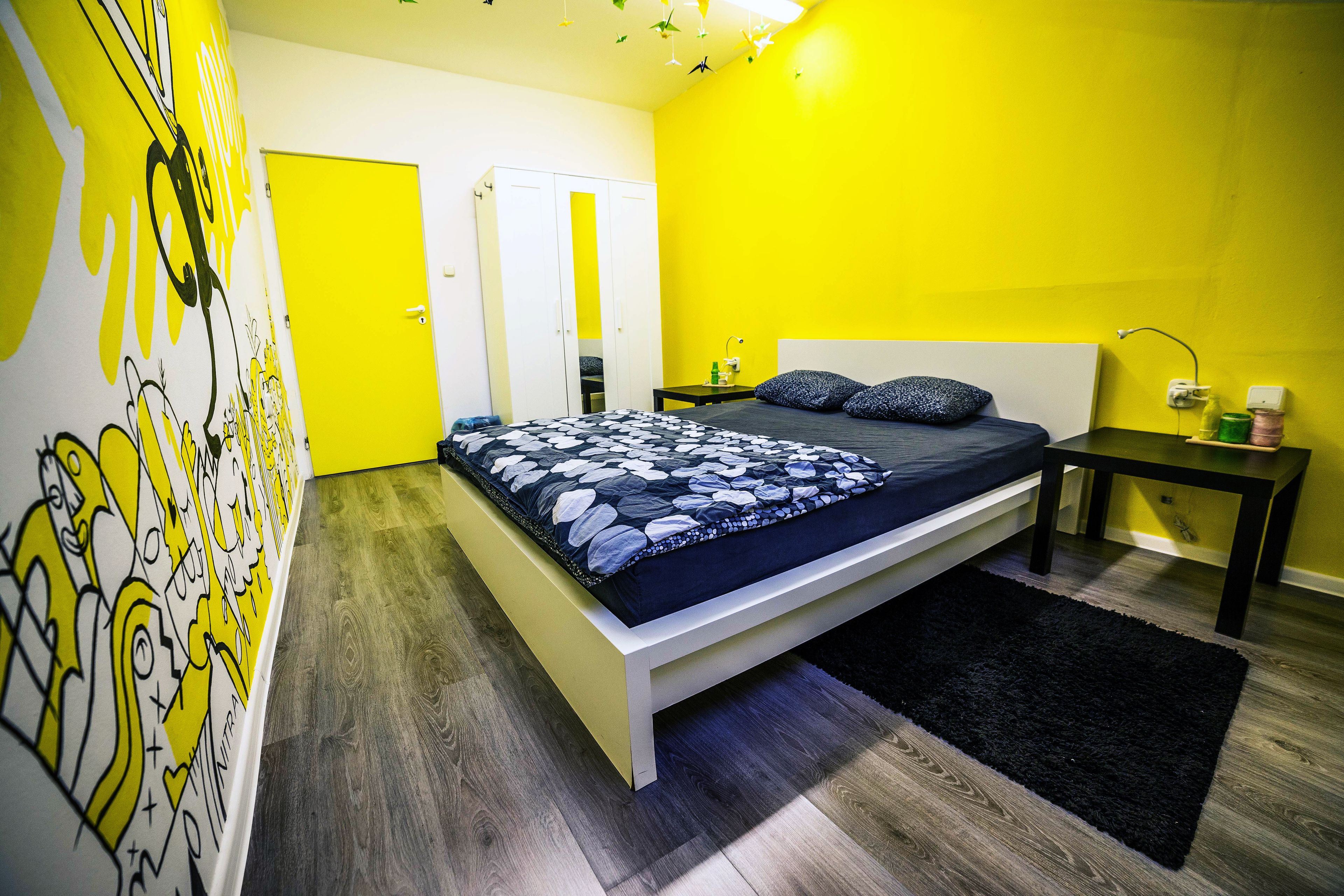 Quarto com cama, mesa de cabeceira, parede com mural e porta.