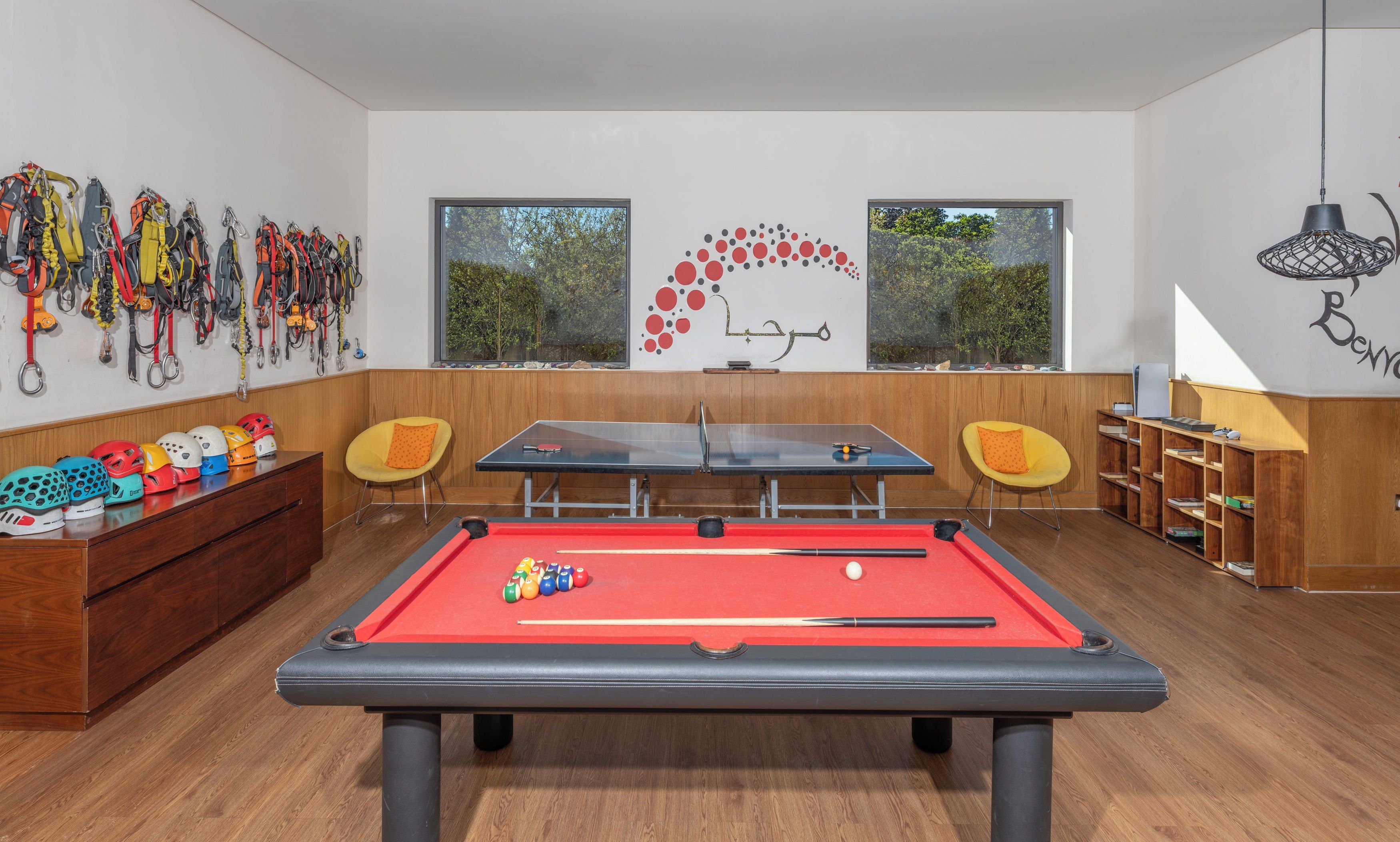Uma mesa de bilhar com superfície de feltro vermelho, rodeada por mobiliário e cadeiras de madeira.