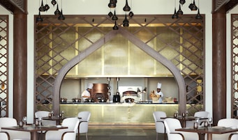 Restaurante com cozinha central, divisória de madeira treliçada e cadeiras brancas.