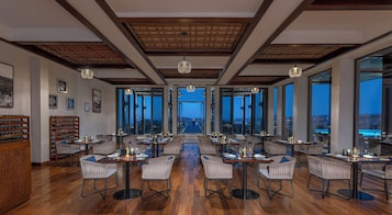 Restaurante com mesas alongadas, amplas janelas e vista para a paisagem.