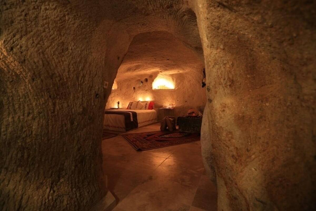 Interior acolhedor e com pouca luz, com uma cama, uma mesinha e um tapete, sugerindo um quarto privado numa estrutura semelhante a uma gruta.