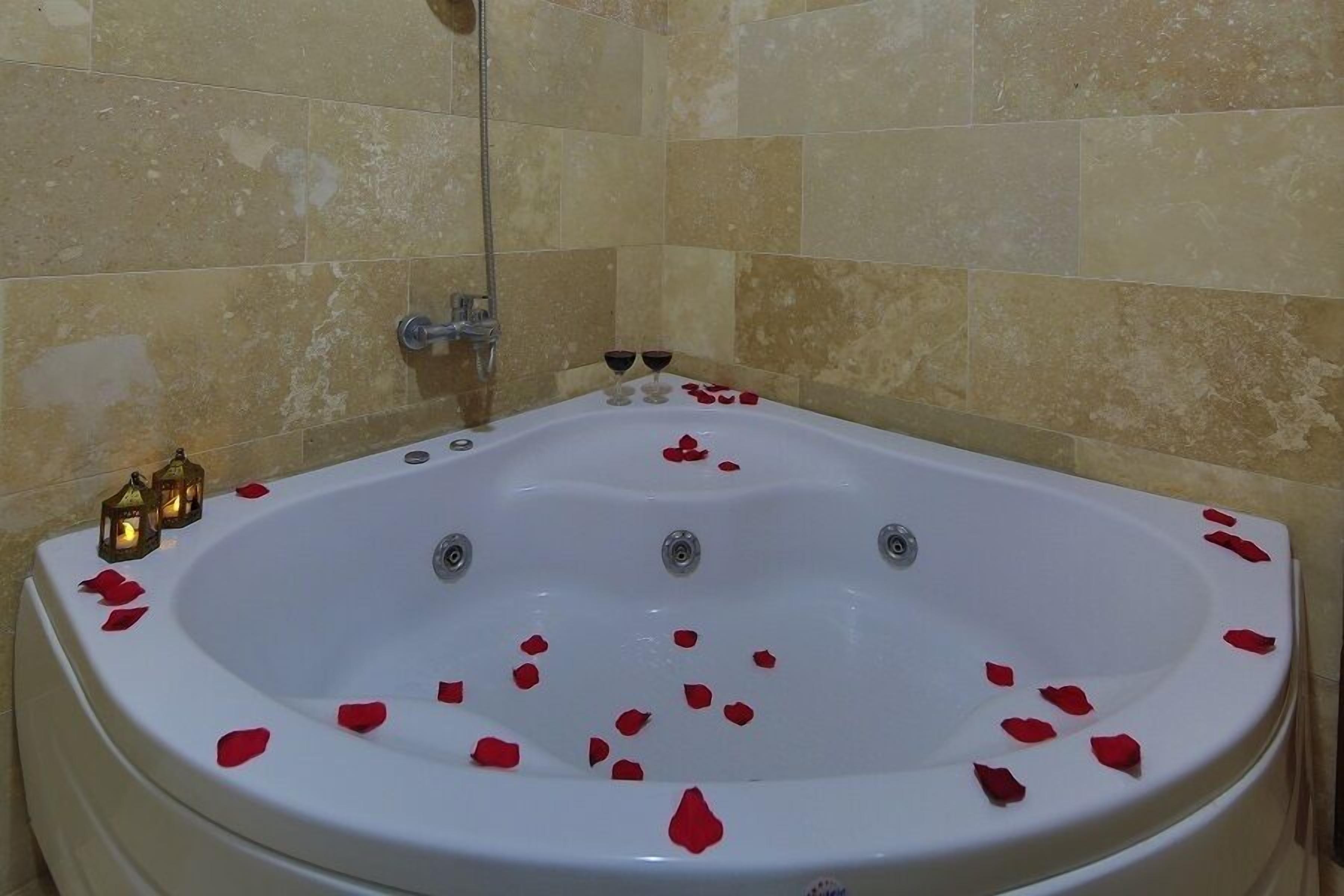 Uma banheira grande e oval, cheia de pétalas de rosas e com duas velas acesas, diante de paredes revestidas com azulejos bege.