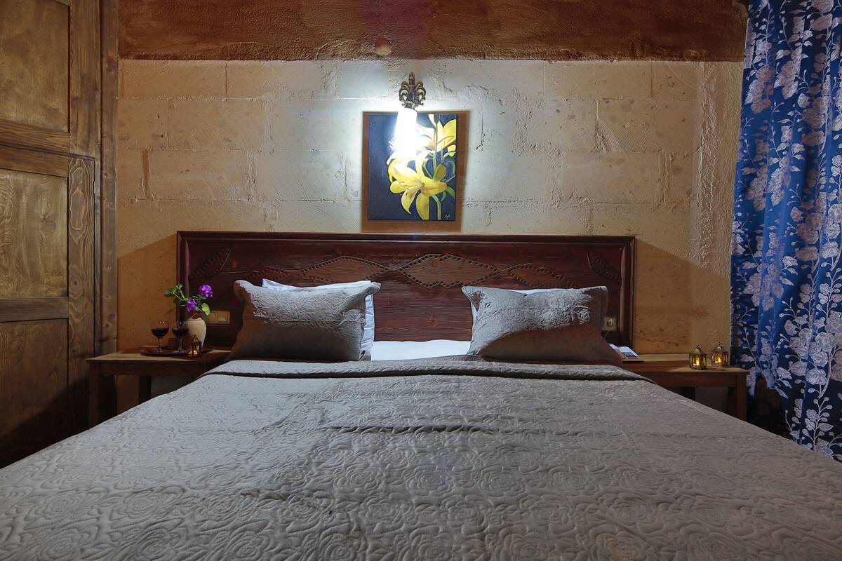 Cama bem arrumada com cabeceira estampada com flores, uma mesa pequena com um vaso e uma vela, e um quadro sobre a cama.