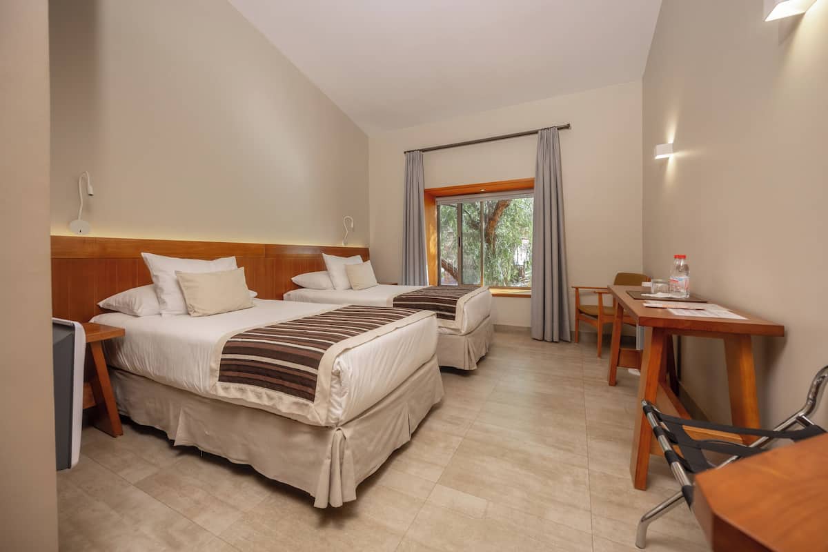 Quarto de hotel com duas camas, uma escrivaninha e uma janela com cortinas.