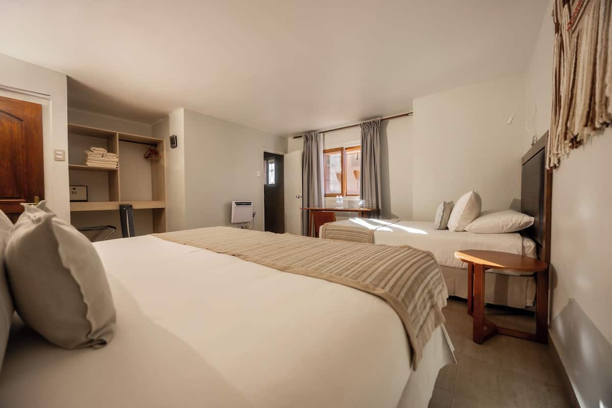 Quarto de hotel com duas camas, um guarda-roupa, uma secretária e uma janela com cortinas.