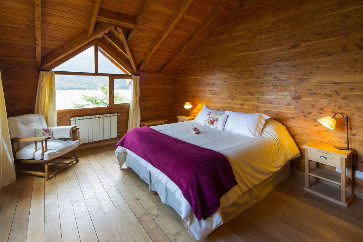 Quarto de cabana de madeira com cama, poltrona de balanço, mesa de cabeceira e janela com vista para um lago.
