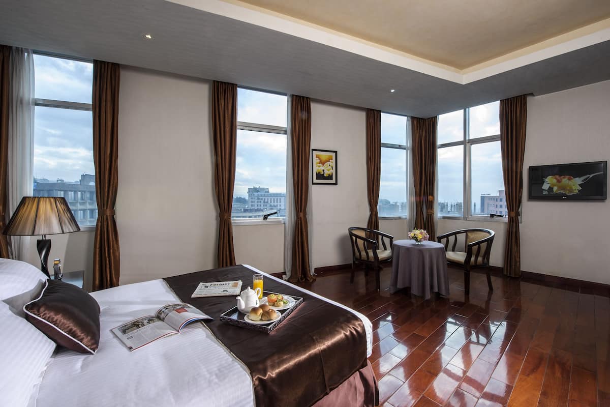 Quarto de hotel com uma cama, uma mesa de jantar e uma cadeira. Há uma janela com cortinas, um abajur e um quadro na parede.
