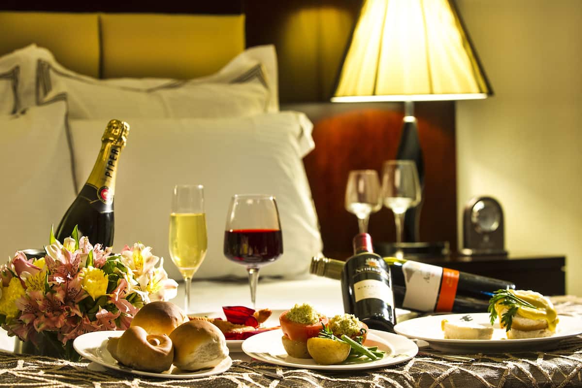 Quarto de hotel com uma mesa de pequeno-almoço bem arrumada, com croissants, uma garrafa de champanhe, copos de vinho e uma peça decorativa floral.