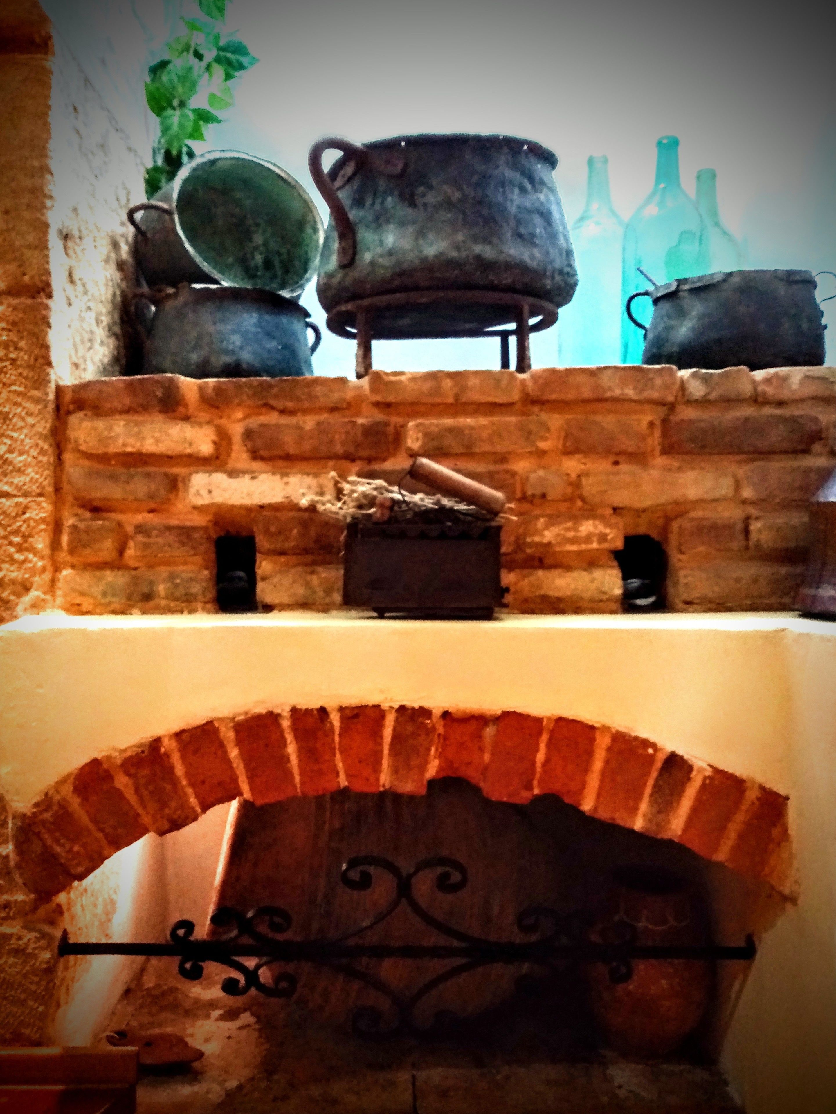 Uma lareira de tijolos com uma grade de metal decorativa e uma prateleira de madeira rústica acima, com várias panelas e frigideiras de metal.