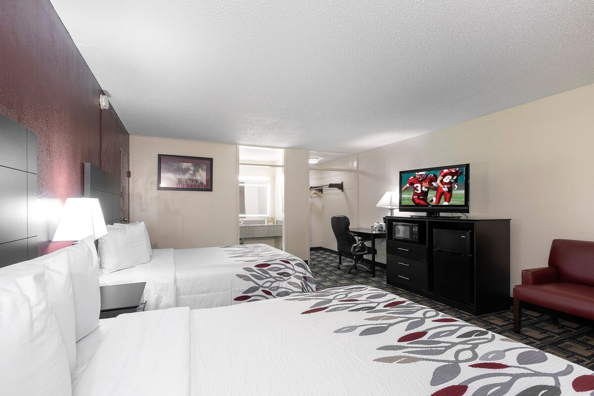 Quarto de hotel com duas camas, uma escrivaninha, uma televisão e uma cadeira vermelha.