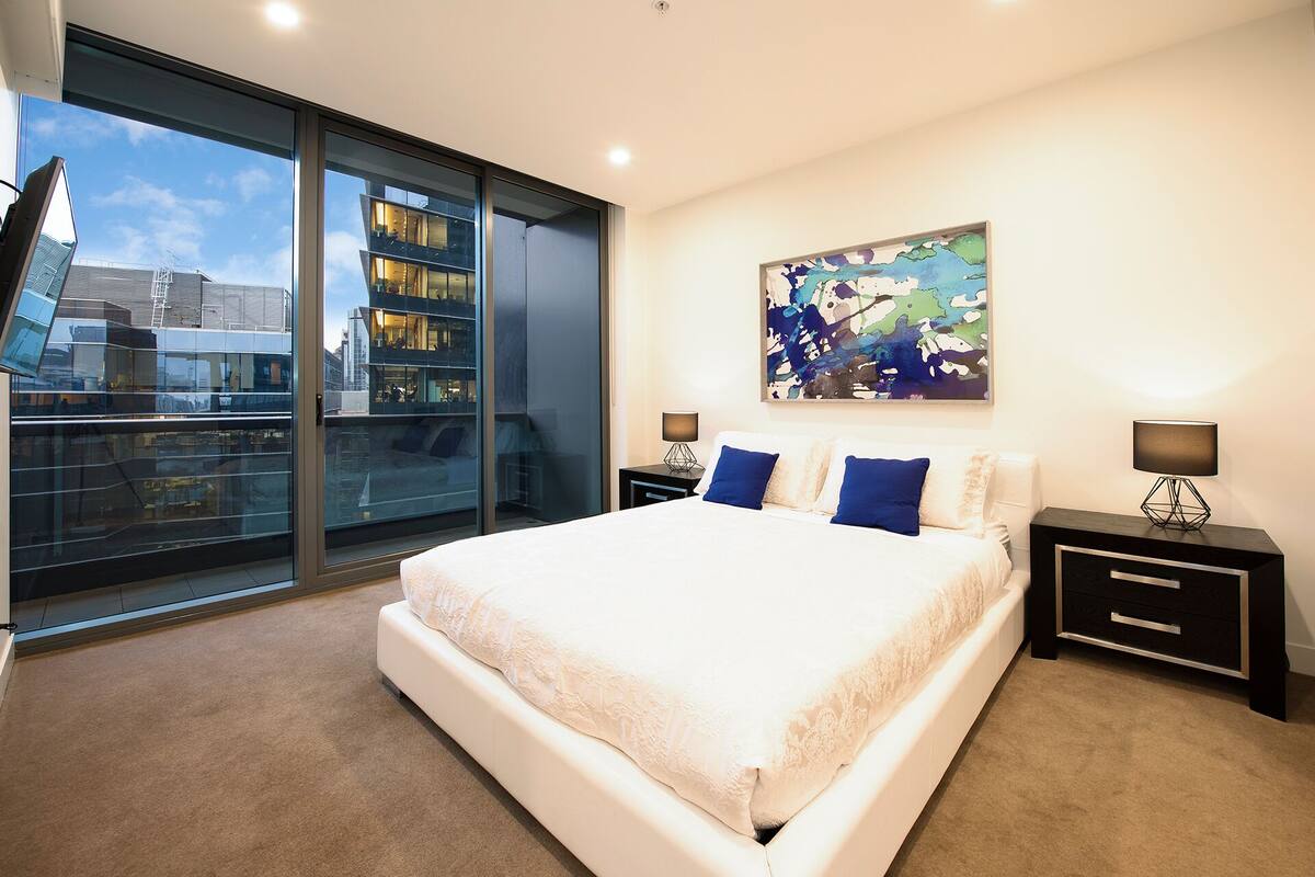Quarto moderno com cama grande, vista da cidade através de janelas do chão ao teto e uma pintura abstrata contemporânea na parede.