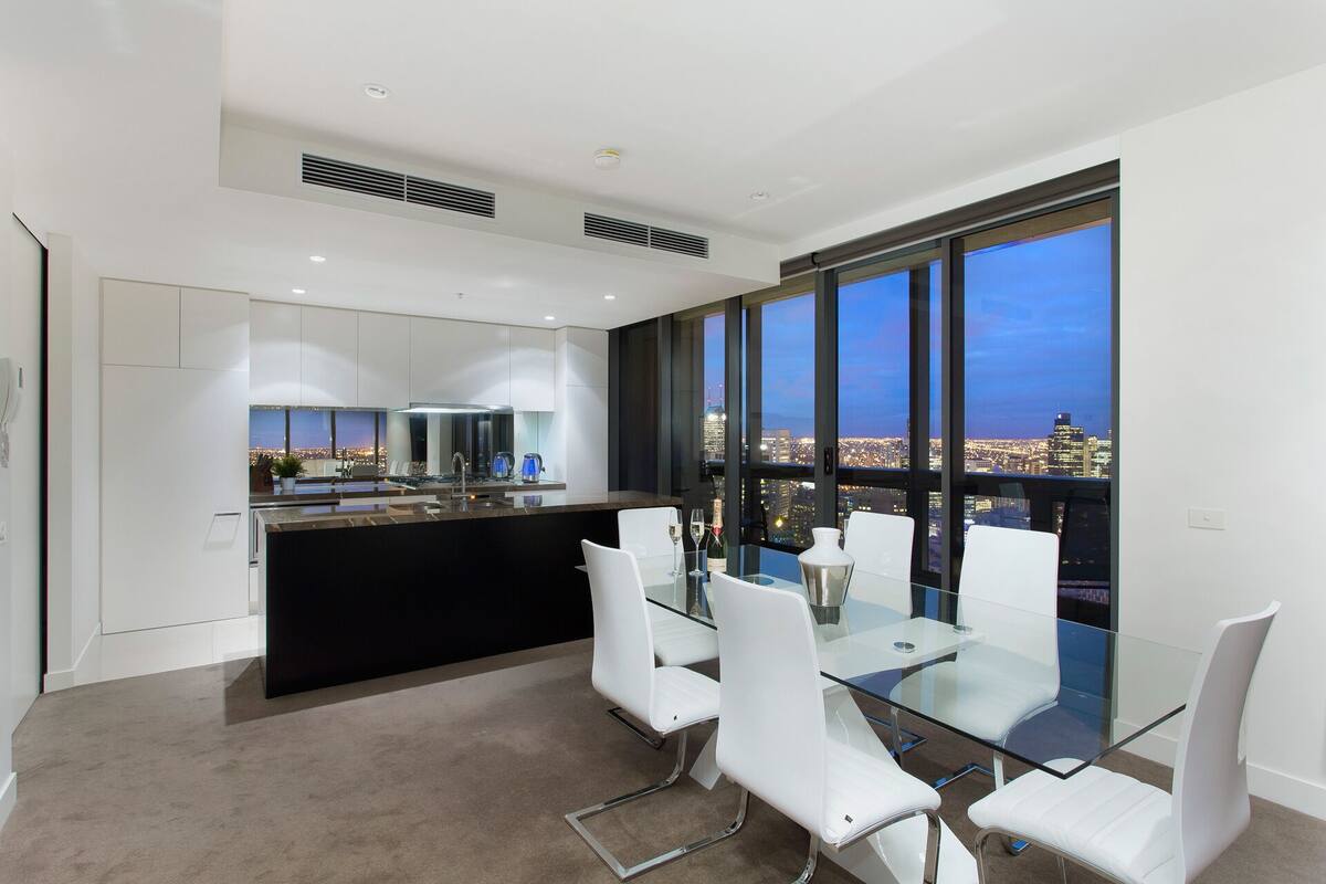 Cozinha moderna com uma ilha grande, sala de jantar com mesa de vidro e cadeiras brancas, e uma janela ampla com vista para a cidade.