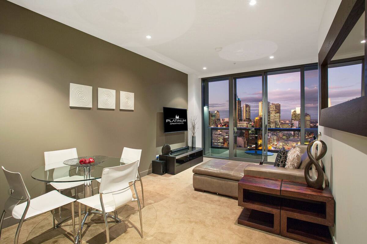 Uma sala de estar moderna com uma mesa de jantar de vidro, cadeiras brancas, um sofá e uma grande janela com vista para a cidade.